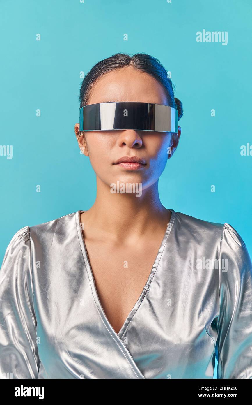 Young trendy ethnic lady with dark hair in modern mirrored VR goggles against blue background ...