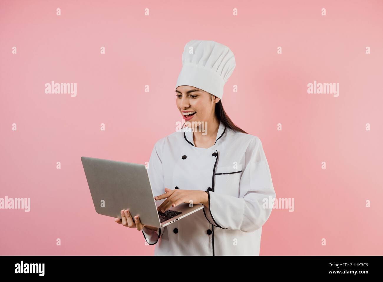 Portrait of young latin woman chef with computer on pink background in ...