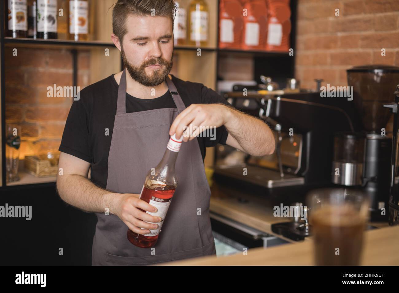 Bearded male barista in apron standing with glass bottle of syrup at ...