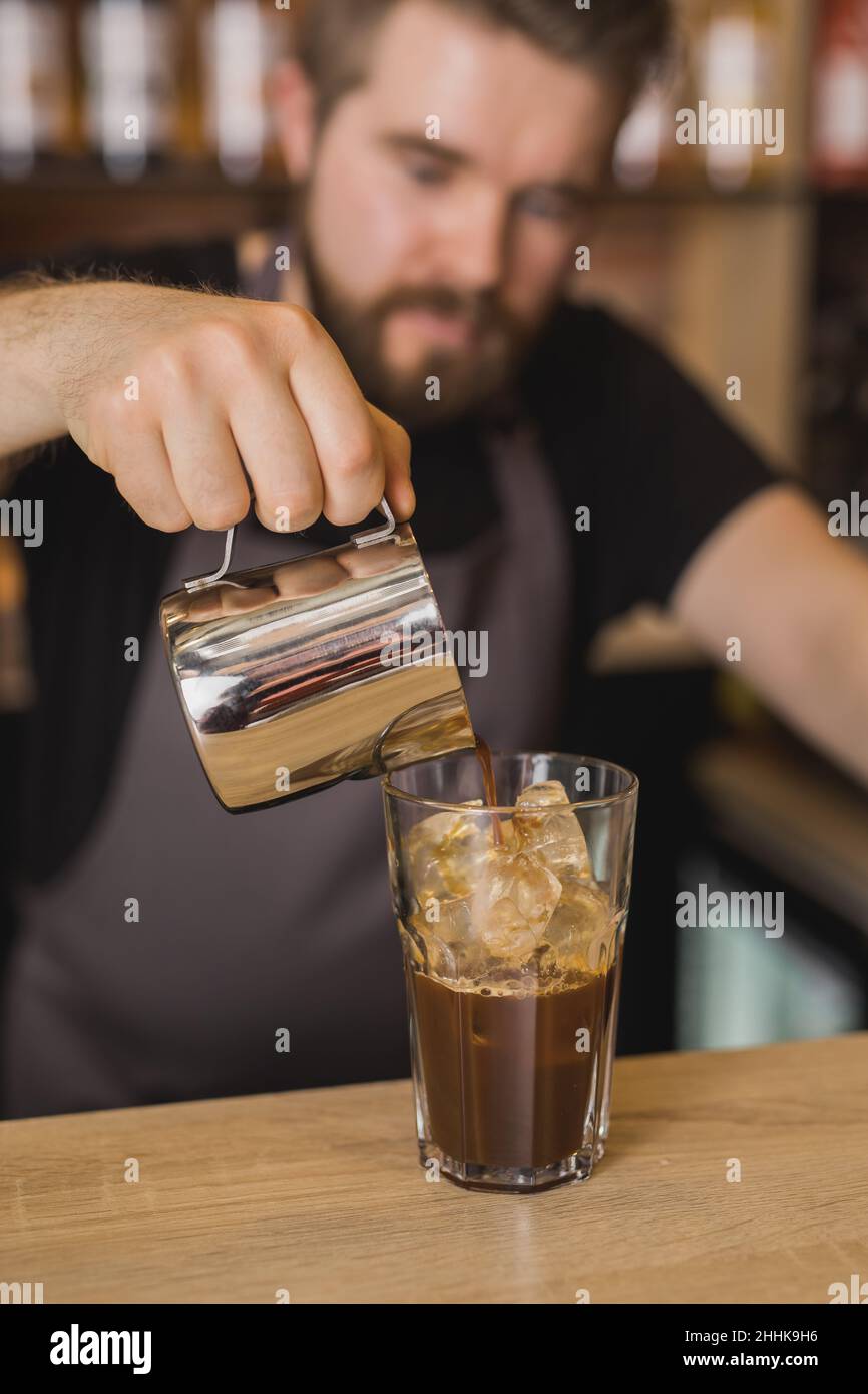 Bearded male barista in apron pouring ingredient from stainless pitcher ...
