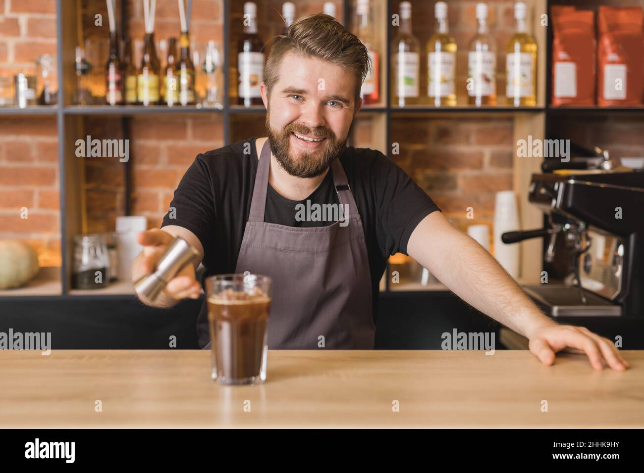 Cheerful bearded male barista in apron pouring syrup from metal jigger ...