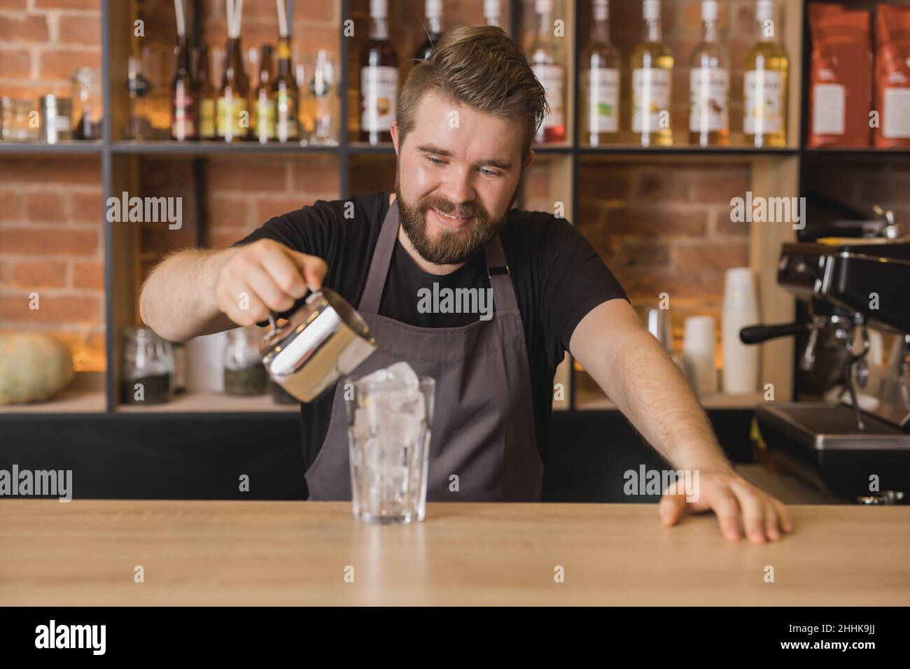 Cheerful bearded male barista in apron pouring ingredient from ...