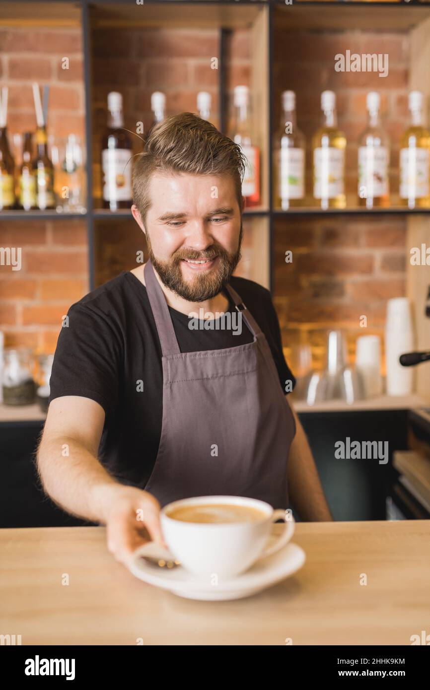 Bearded male barista in apron serving cup of coffee while working at ...