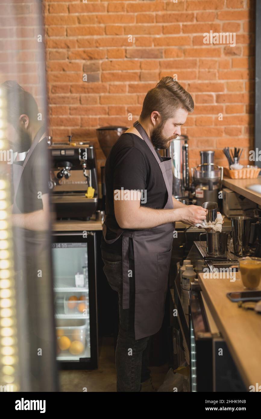 Side view of bearded male barista cleaning pitcher with napkin while ...