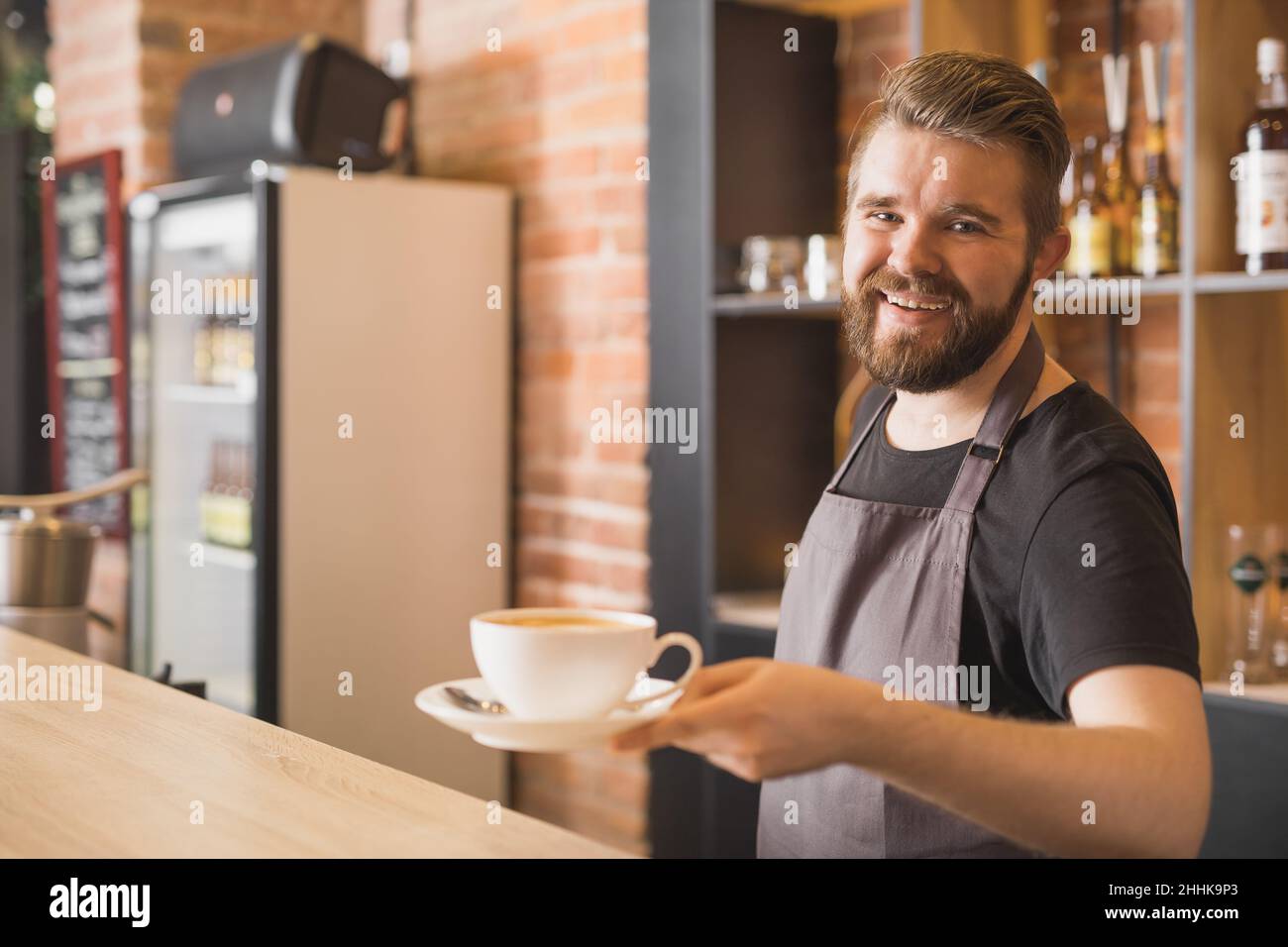 Bearded male barista in apron serving cup of coffee while working at ...