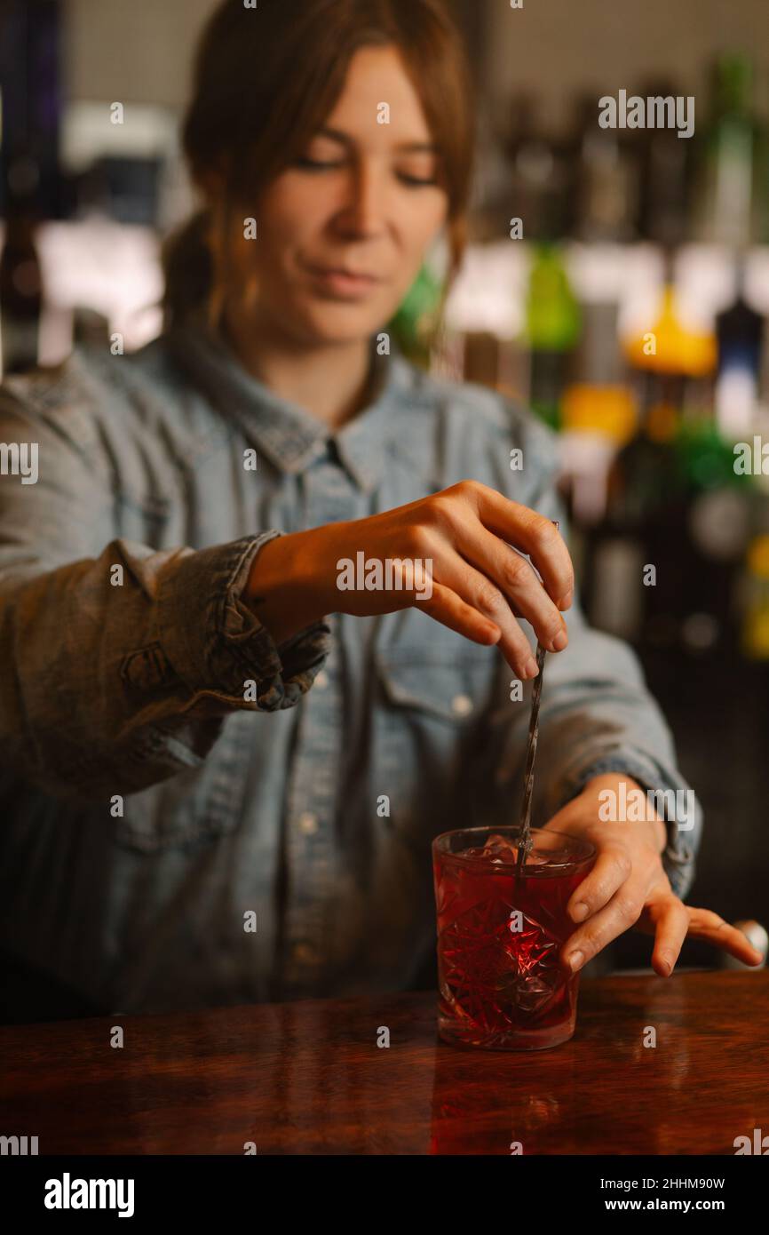 Focused female barkeeper mixing with spoon alcoholic red old fashioned ...