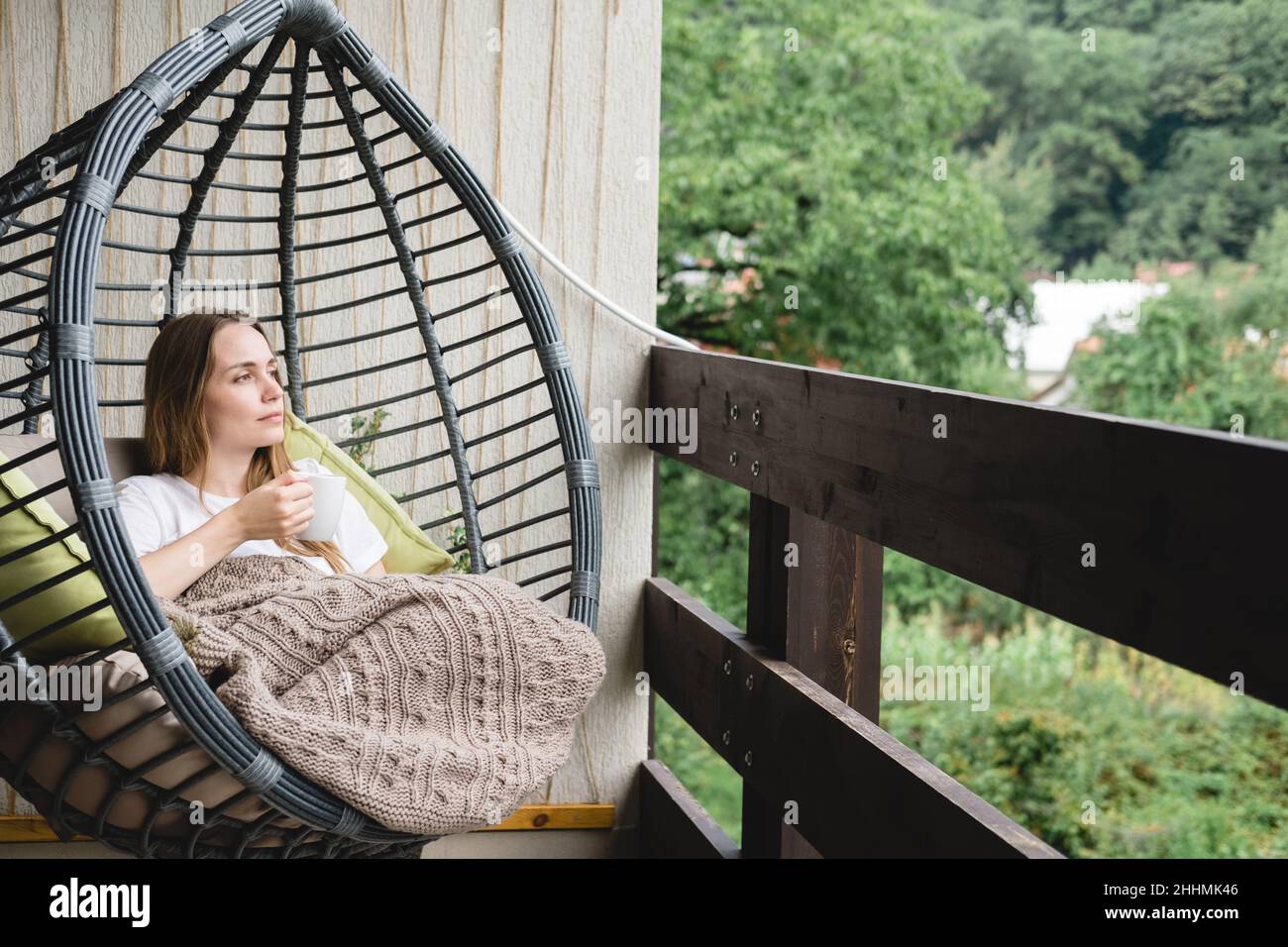 Young woman sitting in a hanging chair with a blanket and a cup of ...