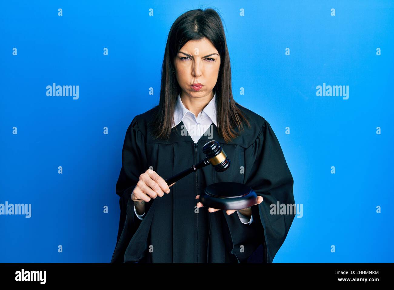 Young hispanic woman using gavel puffing cheeks with funny face. mouth ...