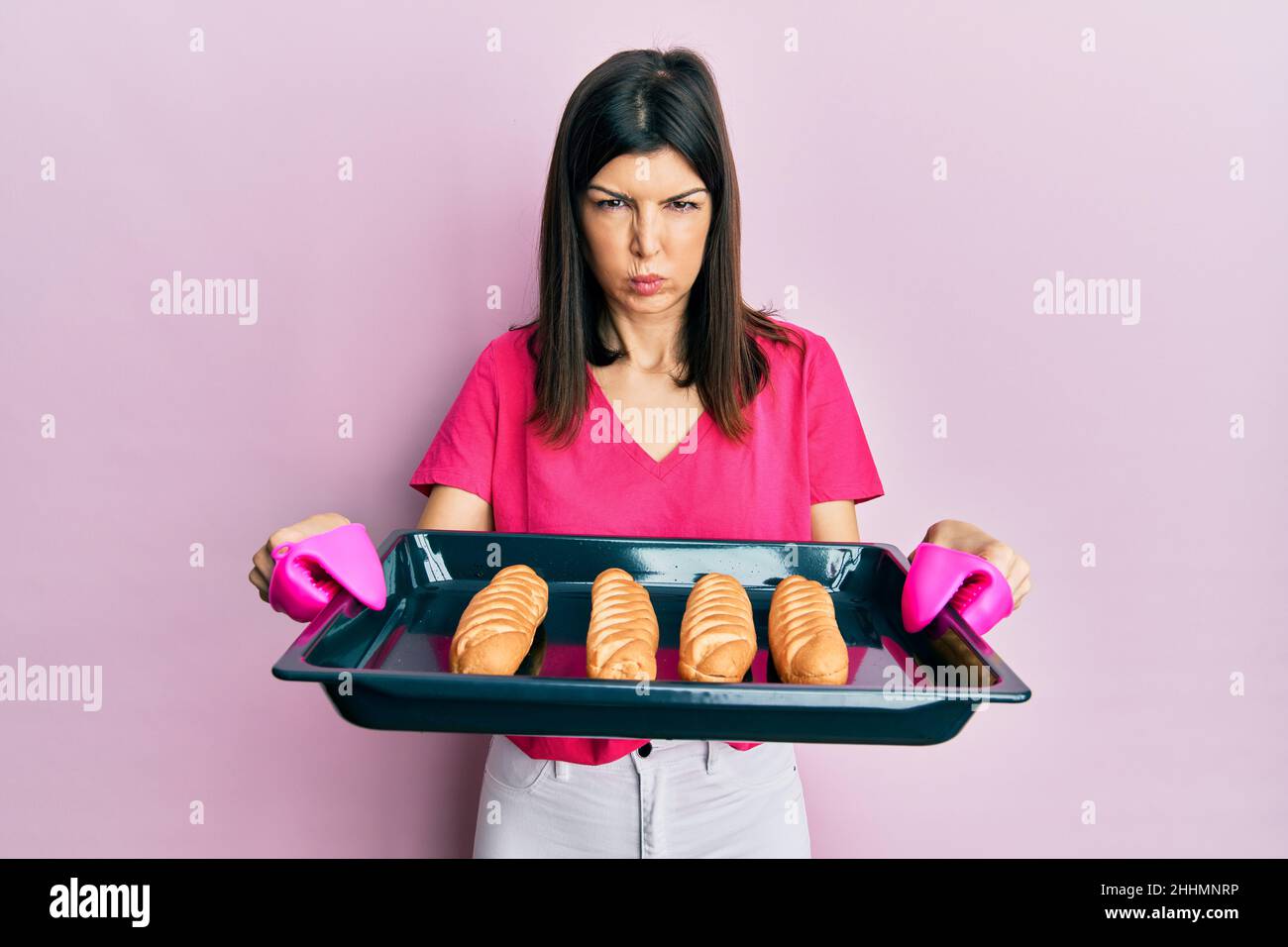 Young hispanic woman holding homemade bread puffing cheeks with funny ...