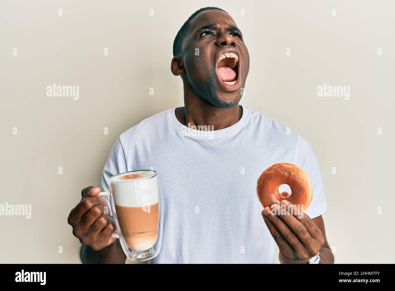 Young african american man eating doughnut and drinking coffee angry ...