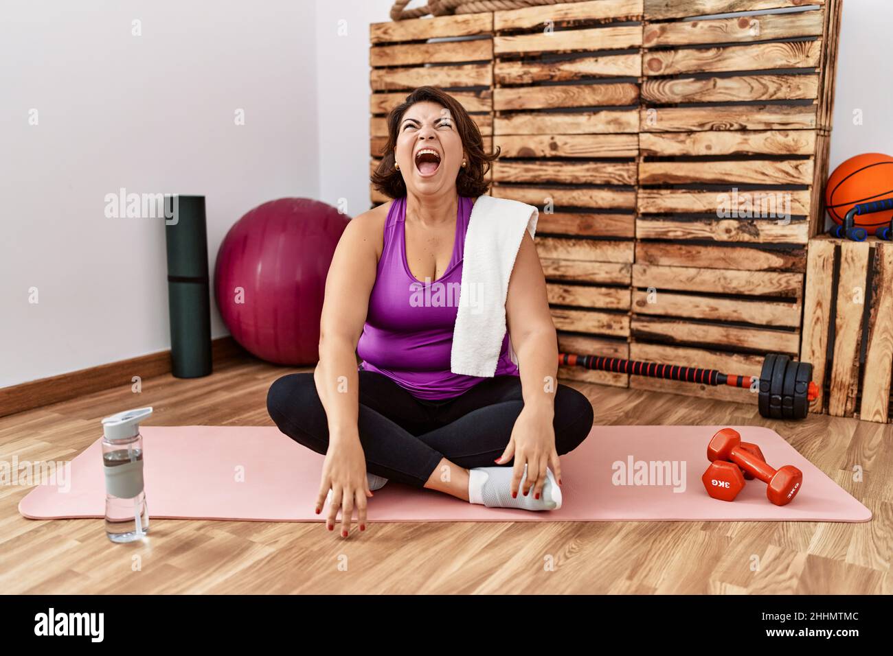 Middle age hispanic woman sitting on training mat at the gym angry and mad screaming frustrated ...