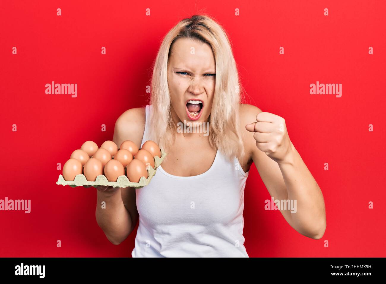 Beautiful caucasian blonde woman holding tray of fresh eggs annoyed and ...