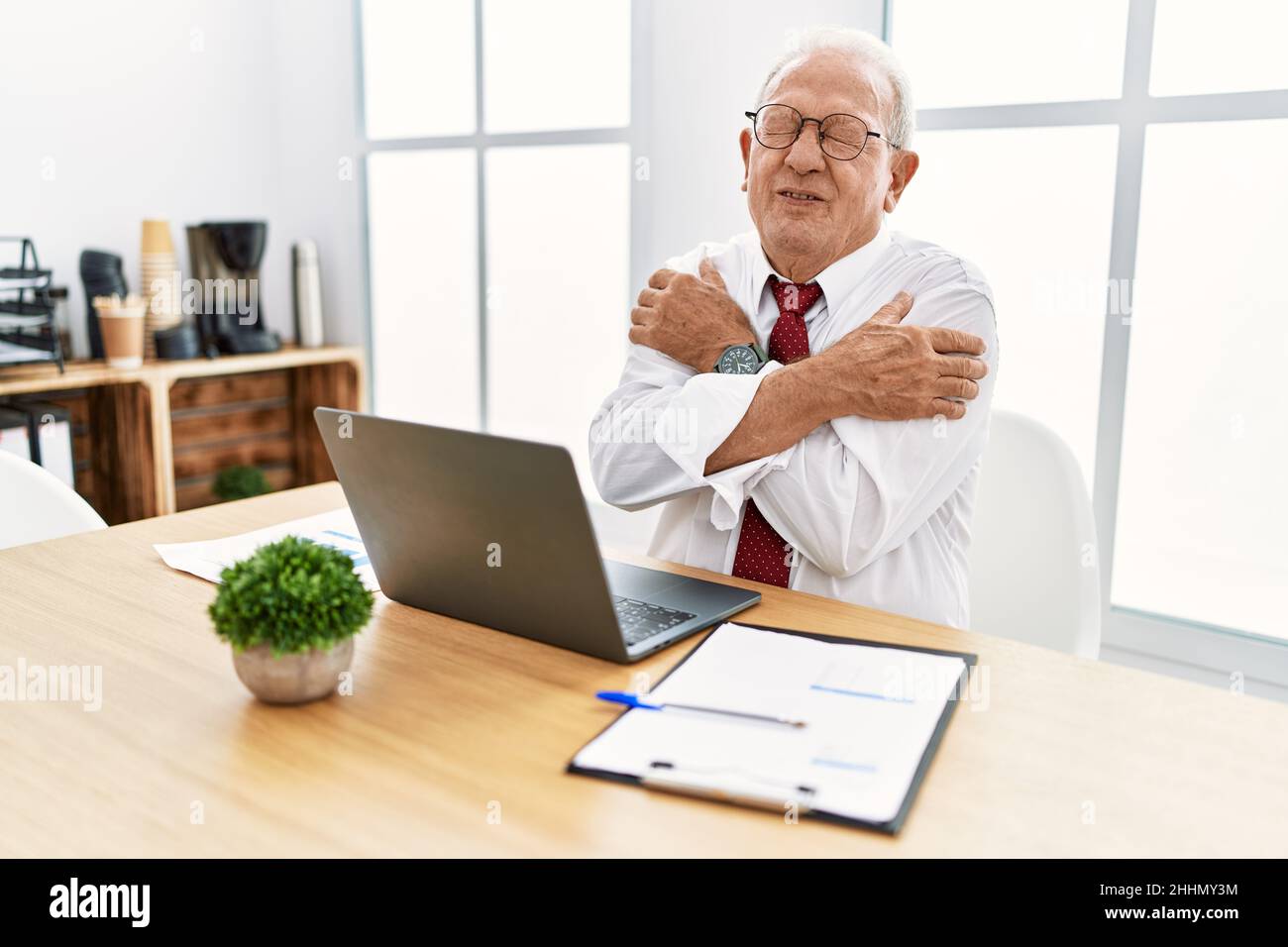 Senior man working at the office using computer laptop hugging oneself ...
