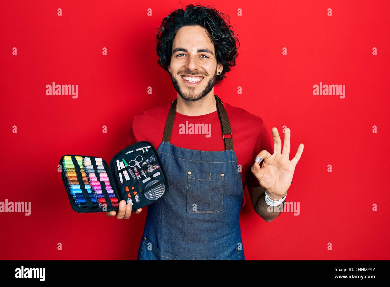 Handsome hispanic man wearing apron holding sew kit doing ok sign with ...