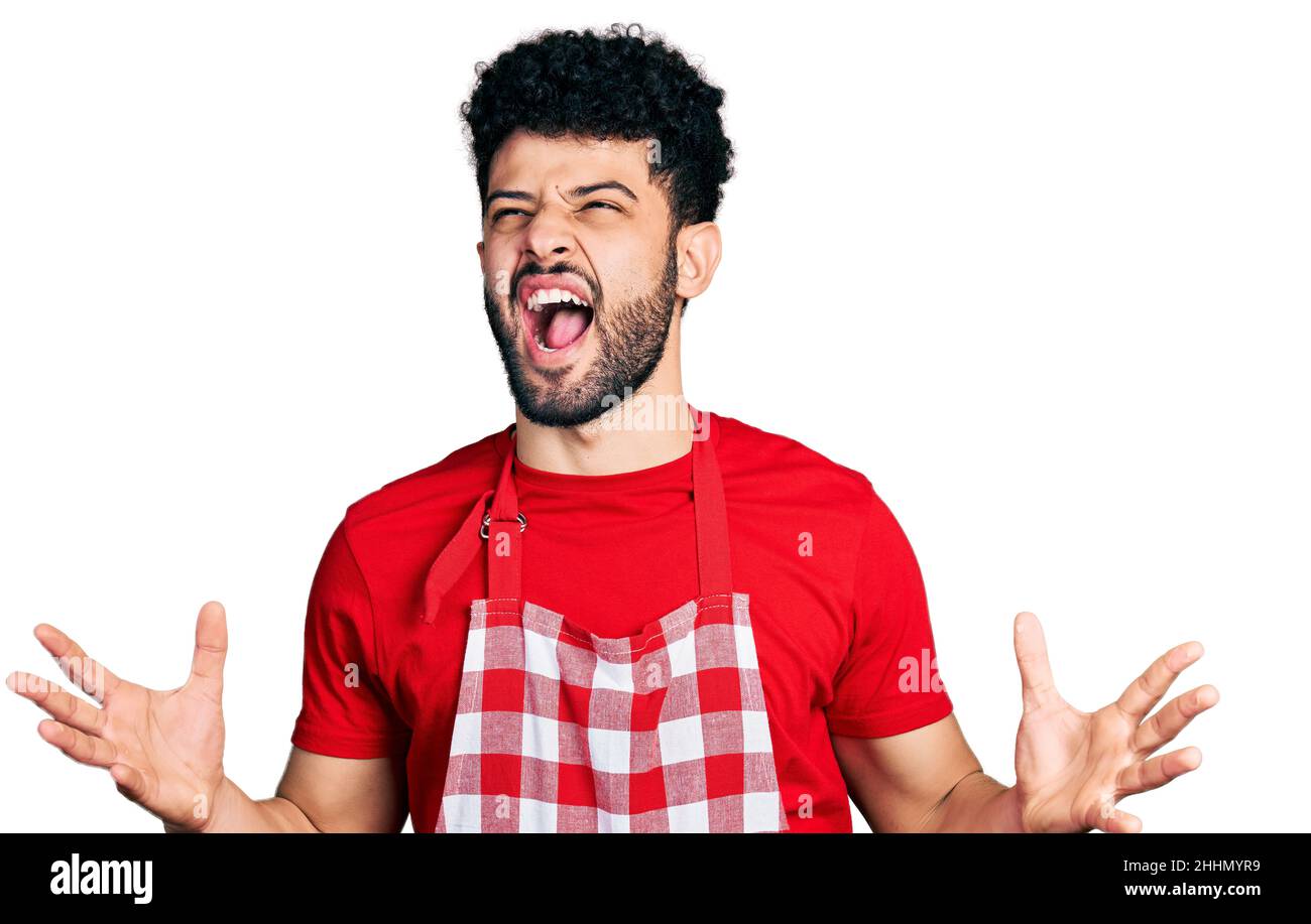 Young arab man with beard wearing cook apron crazy and mad shouting and ...
