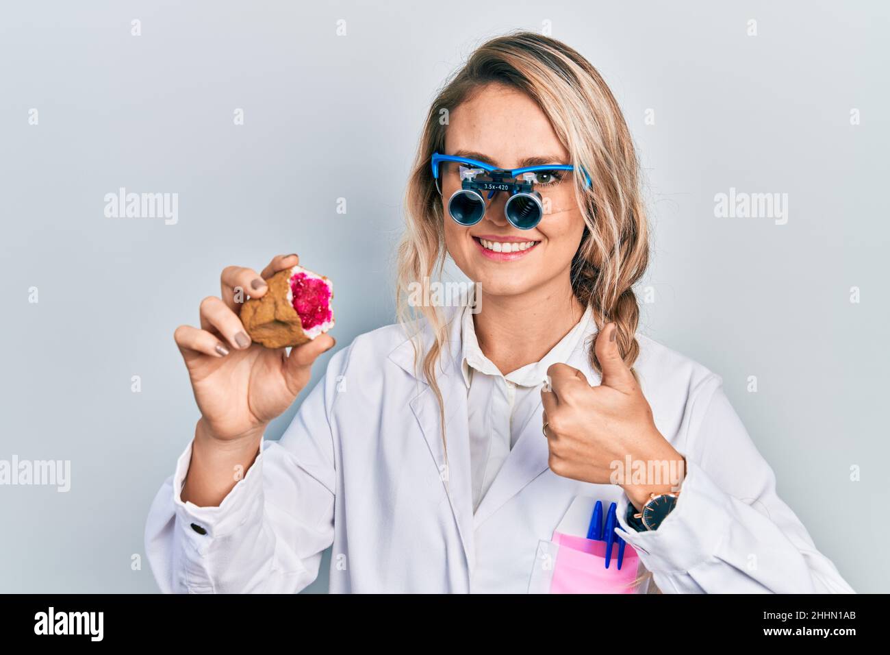 Beautiful young blonde woman holding geode stone wearing magnifier ...