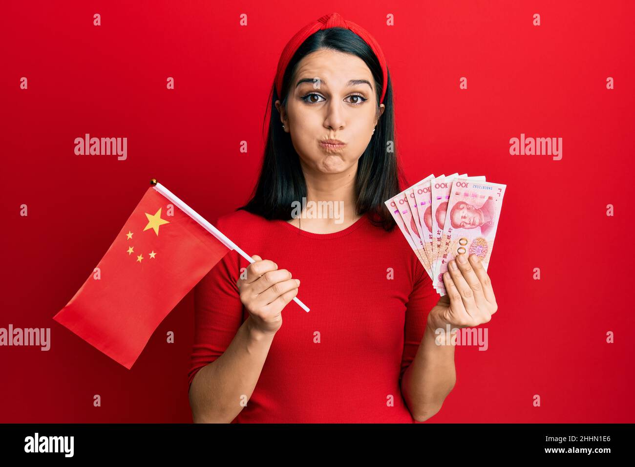 Young hispanic woman holding china flag and yuan banknotes puffing ...
