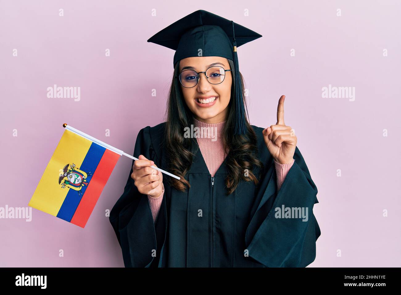 Young hispanic woman wearing graduation uniform holding ecuador flag ...