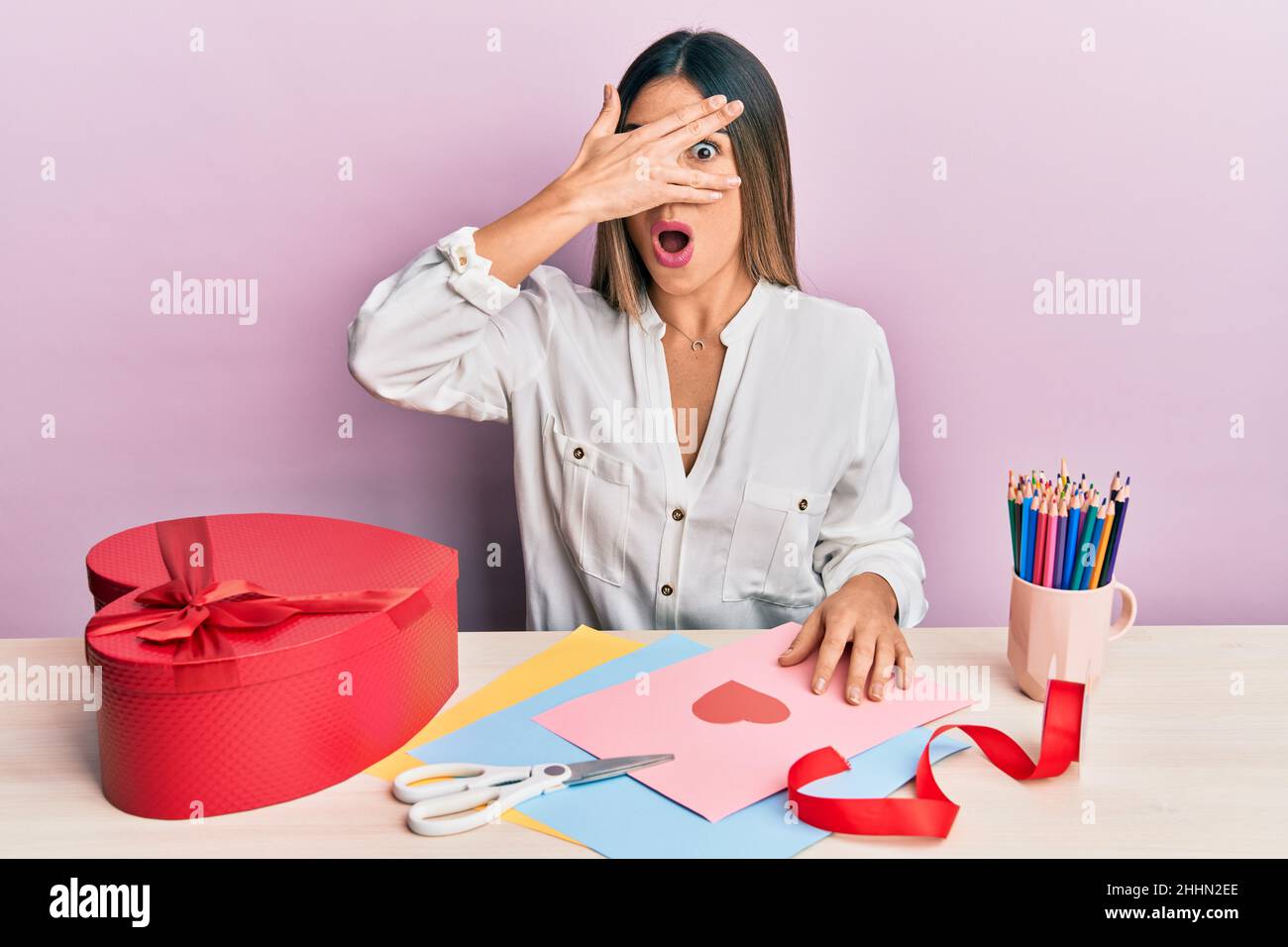 Young hispanic woman making valentine gift sitting on the table peeking ...