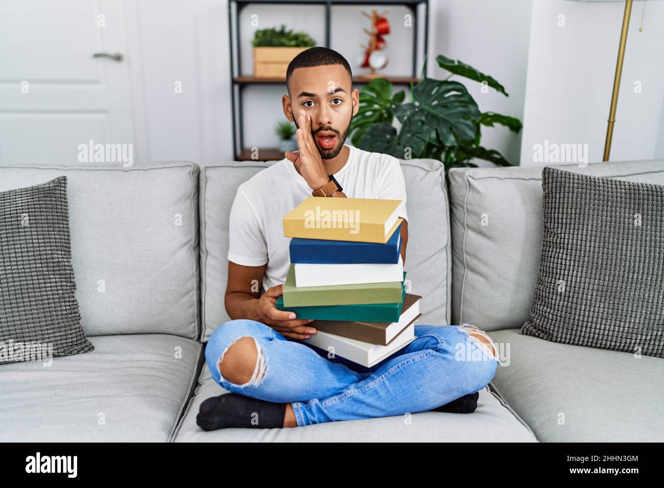 African american young man holding a pile of books sitting on the sofa ...