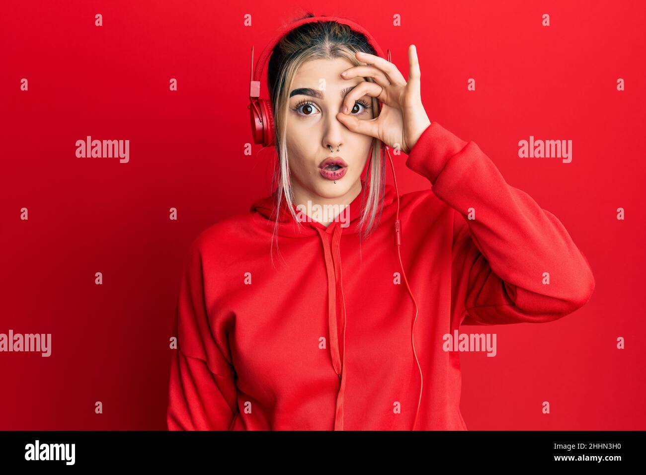 Young modern girl wearing gym clothes and using headphones doing ok gesture shocked with ...