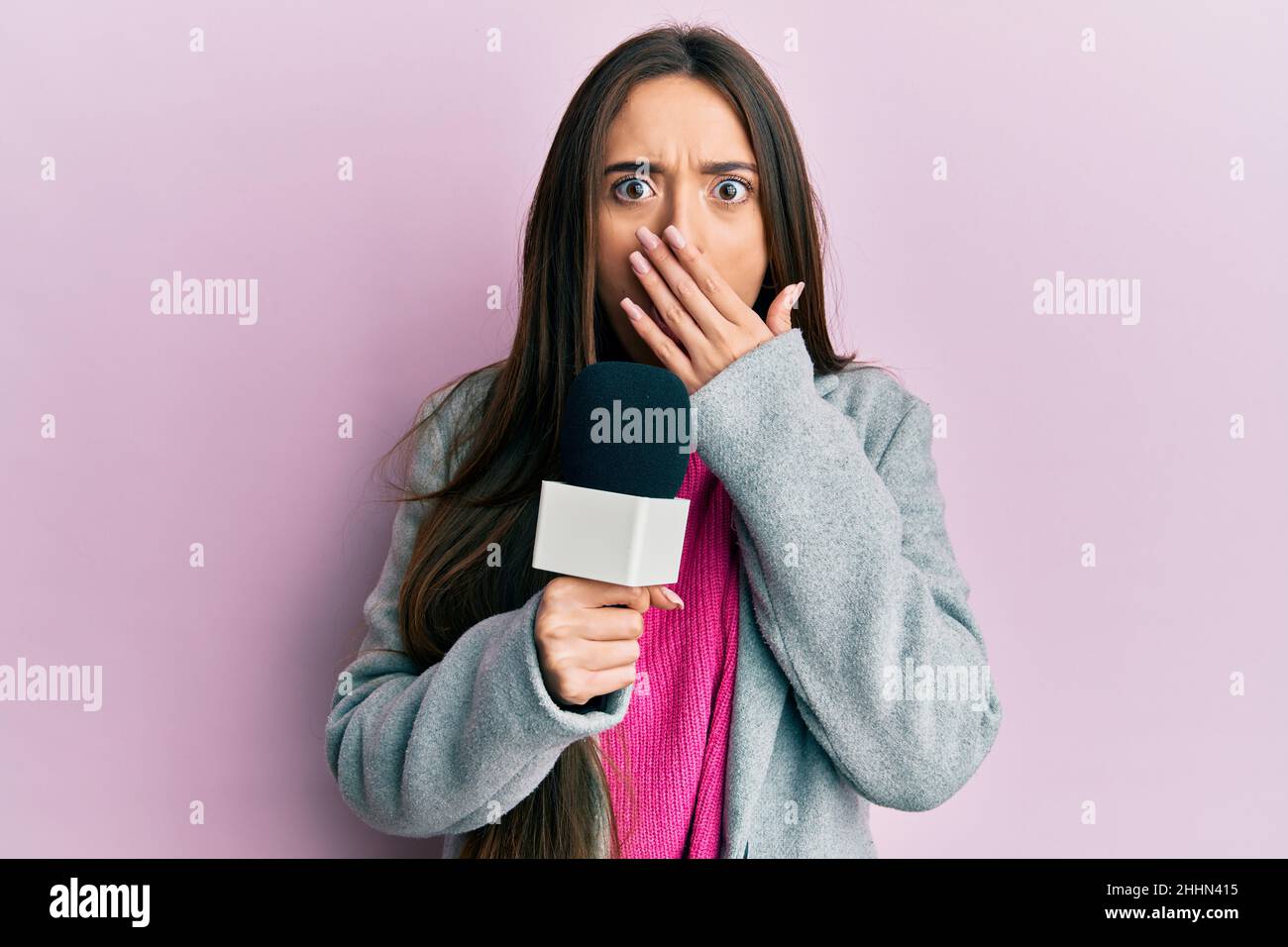 Young hispanic girl holding reporter microphone covering mouth with hand, shocked and afraid for ...