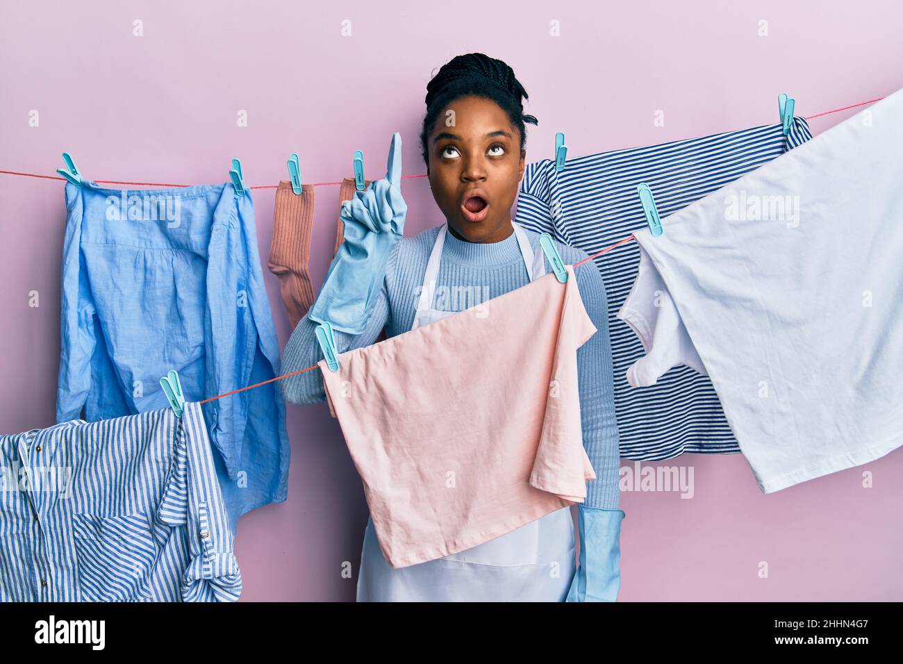 African american woman with braided hair washing clothes at clothesline amazed and surprised ...