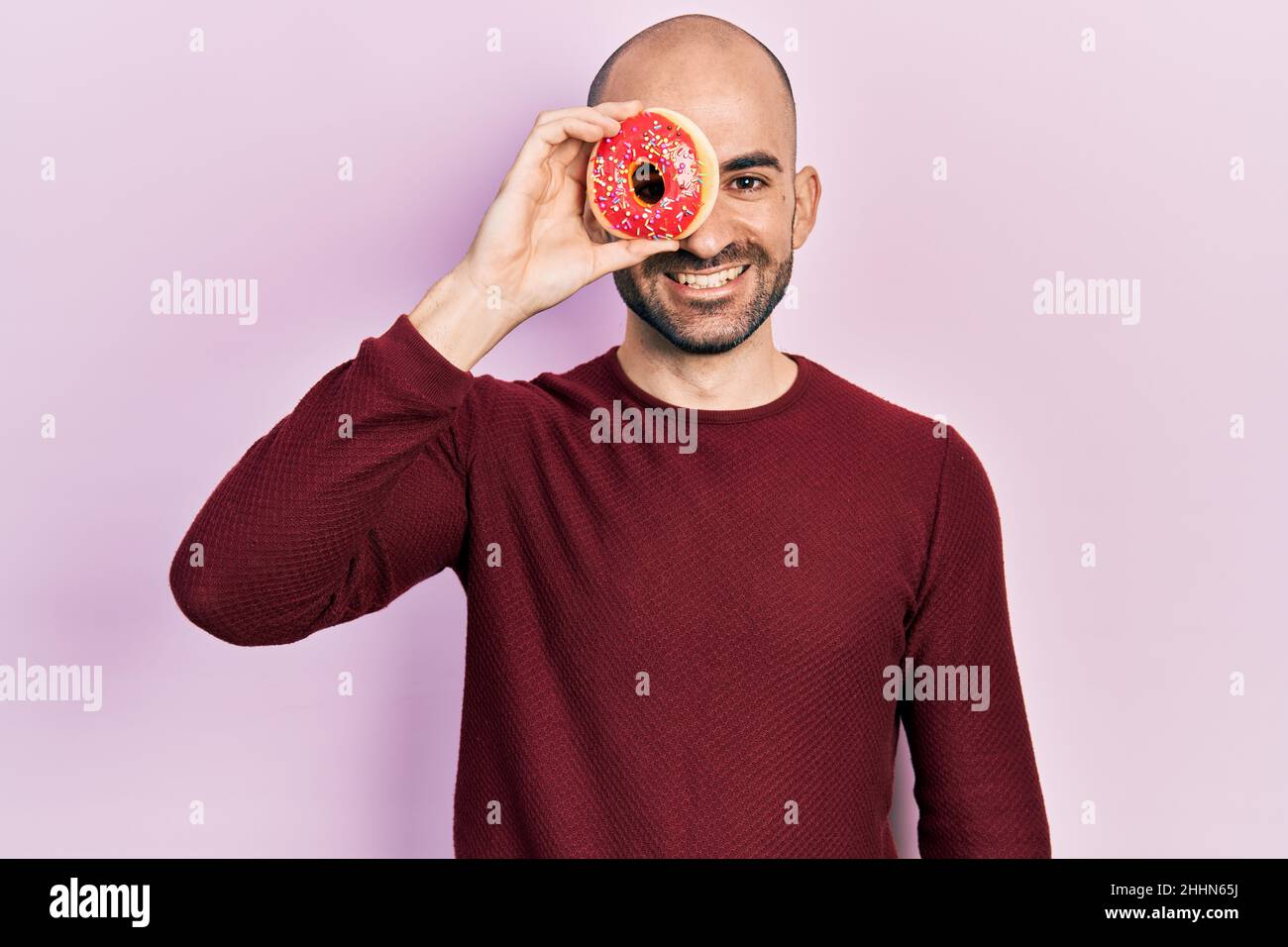 Young bald man holding tasty colorful doughnut on eye looking positive ...
