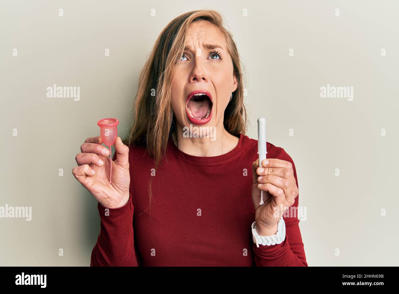 Young blonde woman holding menstrual cup and tampon angry and mad ...