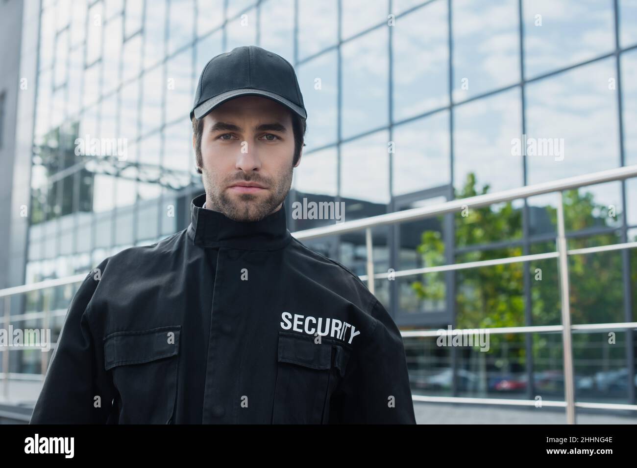 young security man in black uniform looking at camera near building ...