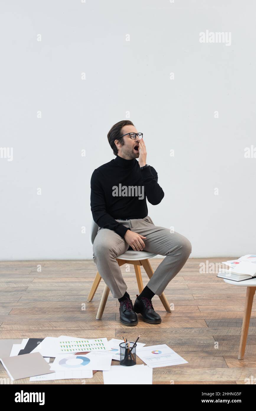 sleepy man yawning on chair near documents on floor and notebooks on ...