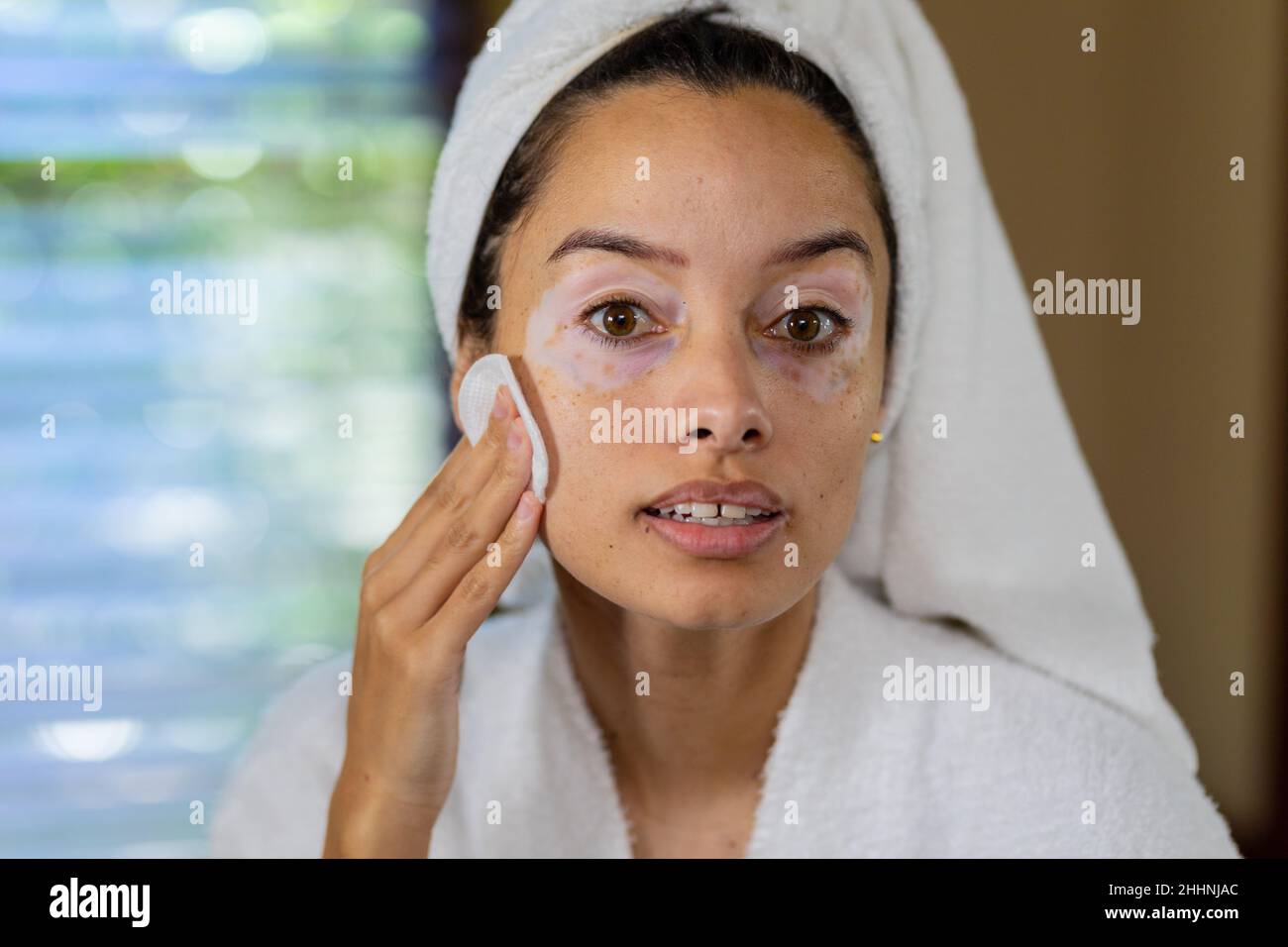 Closeup portrait of biracial young woman in bathrobe applying cream on ...