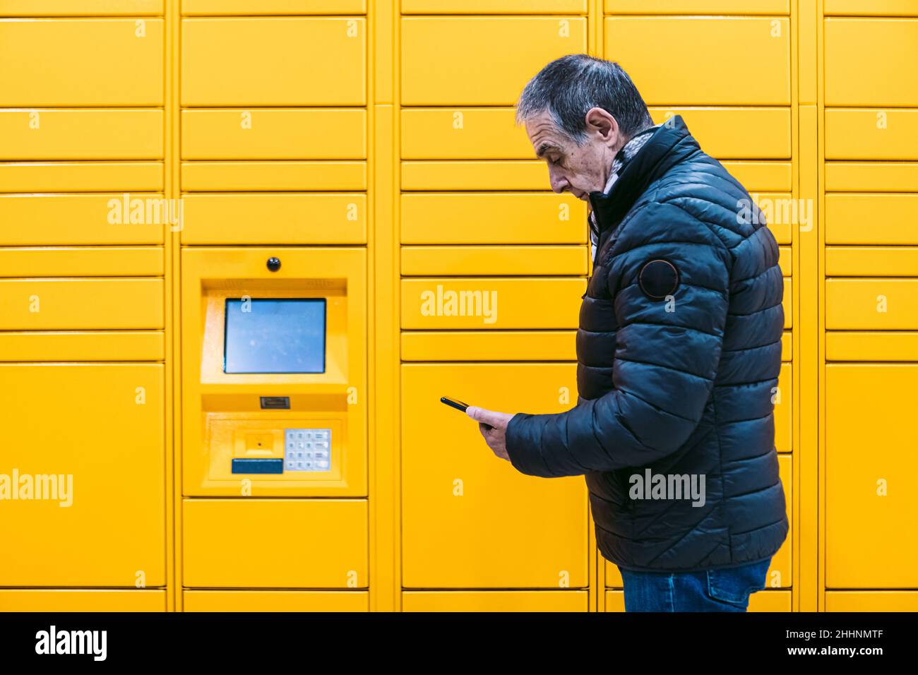 Man with gray coat looks at his cell phone to pick up a package from the yellow locker ...