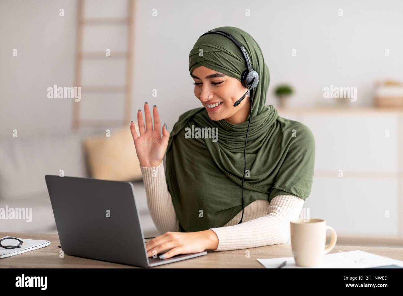 Cheerful young Arab woman in hijab and headphones making video call on ...
