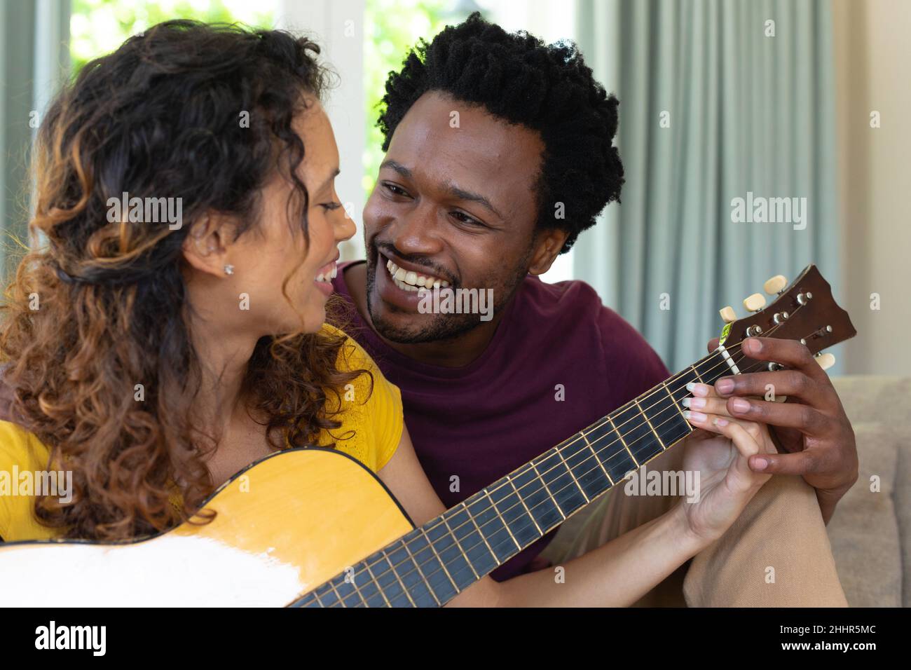 Happy romantic young multiracial couple playing guitar together at home ...