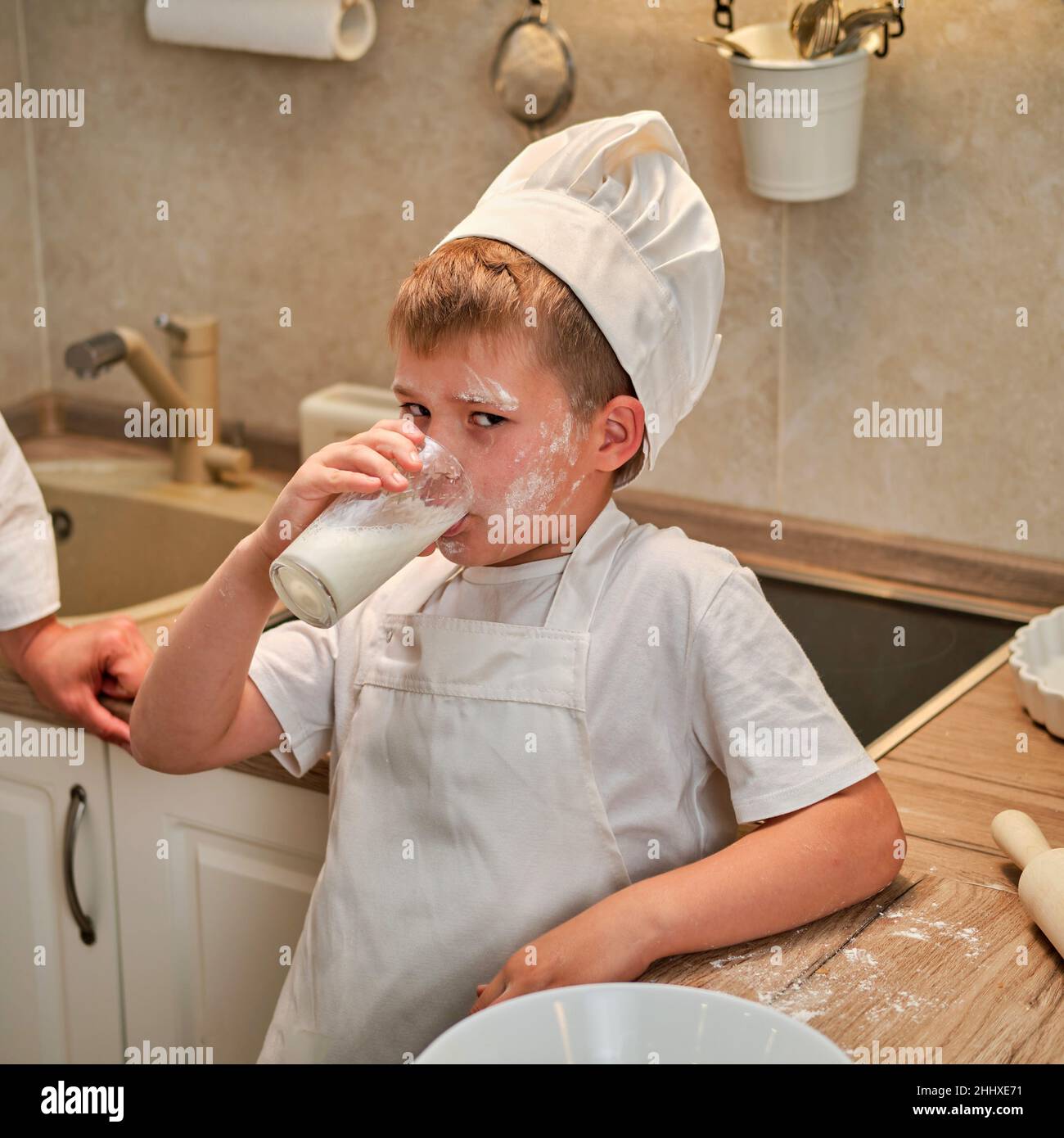 A boy in chef clothes drinks milk from a glass while cooking an apple ...