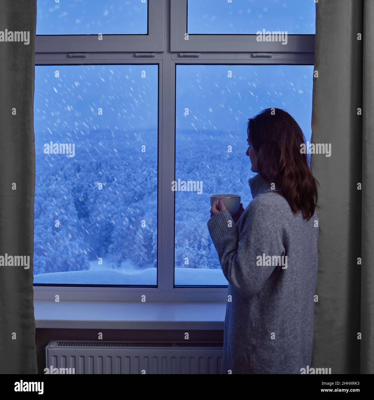 A woman stands at a winter evening window with trees in the snow. A ...