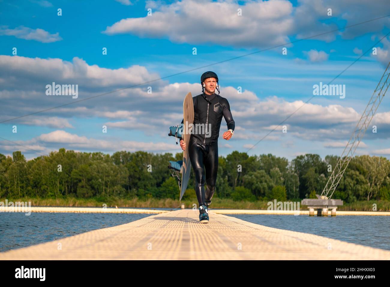 Confident man in wetsuit with wakeboard walking on floating bridge ...