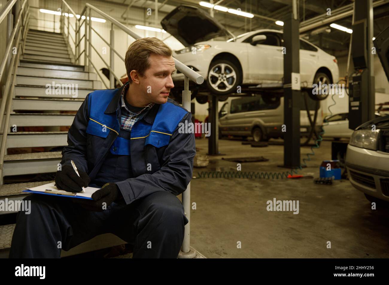 Caucasian mechanic sitting on stairs in the repair shop against cars ...