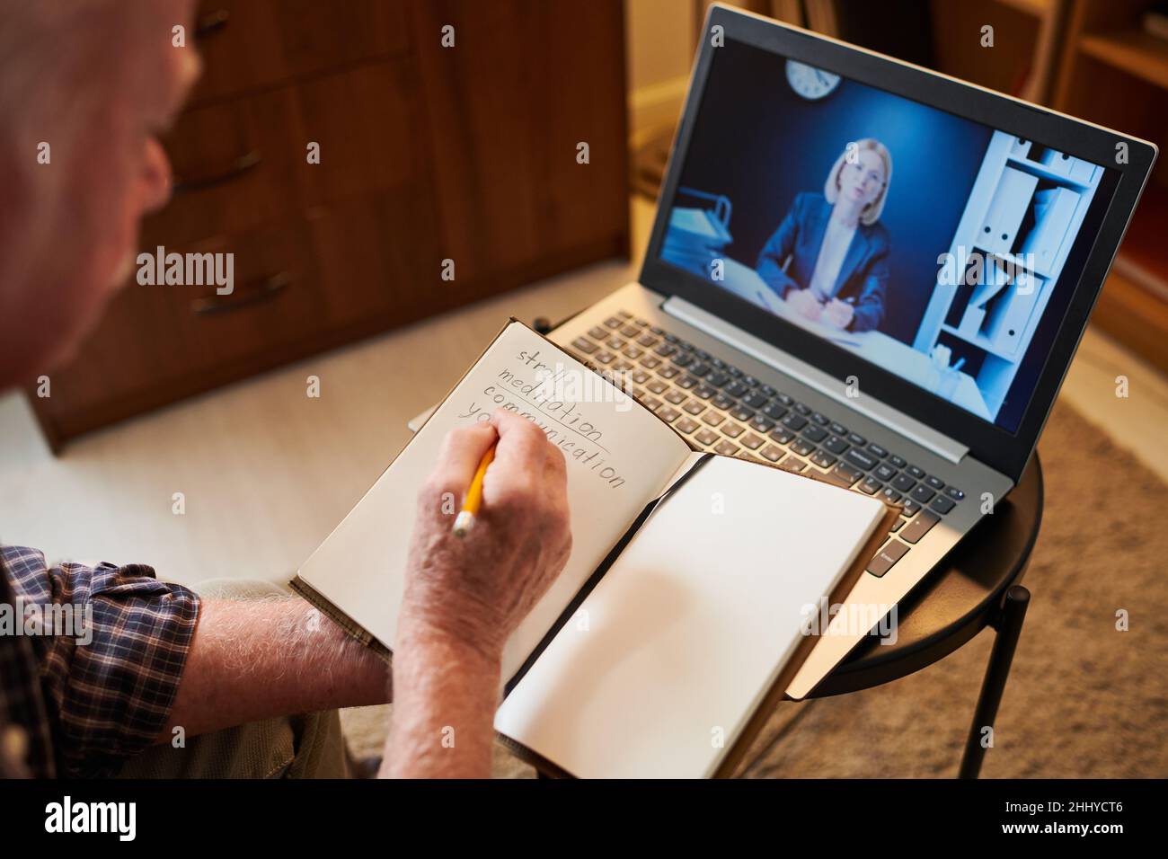 Old man making notes in notebook while sitting in front of laptop ...