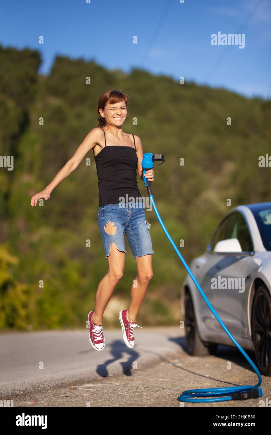 Beautiful young girl jumping next to an electric car. Holding blue ...