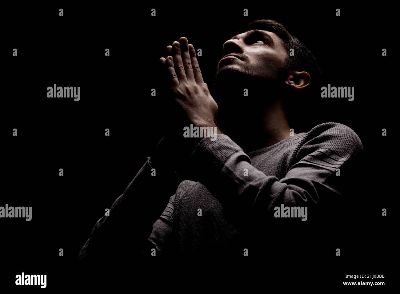 Headshot of serious confident young man praying in dark room. Lit from ...