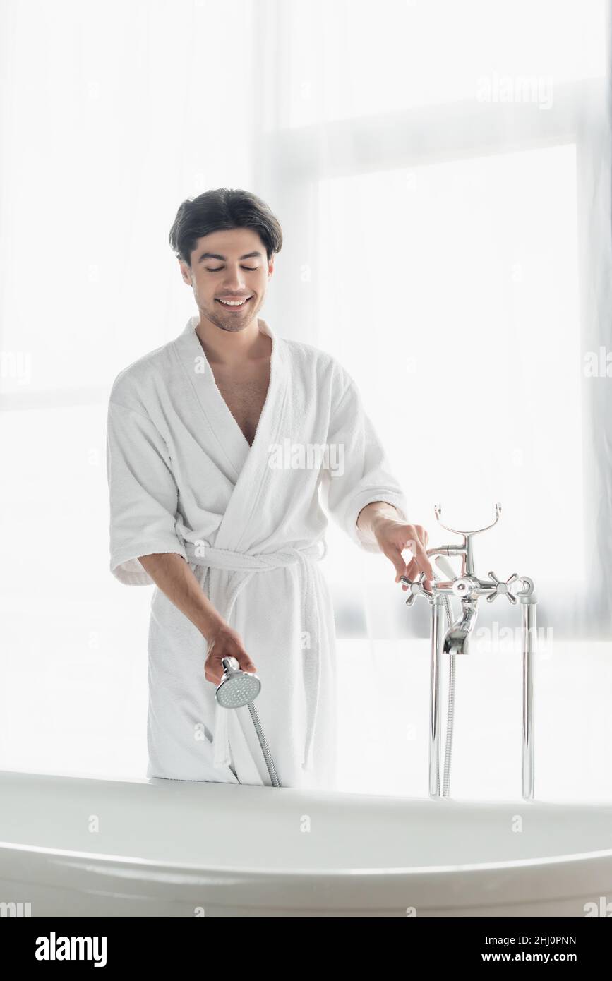 smiling man in white bathrobe holding shower head while opening faucet ...