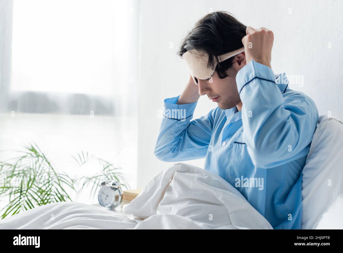 man in blue pajamas sitting on bed in morning and taking off sleep mask ...