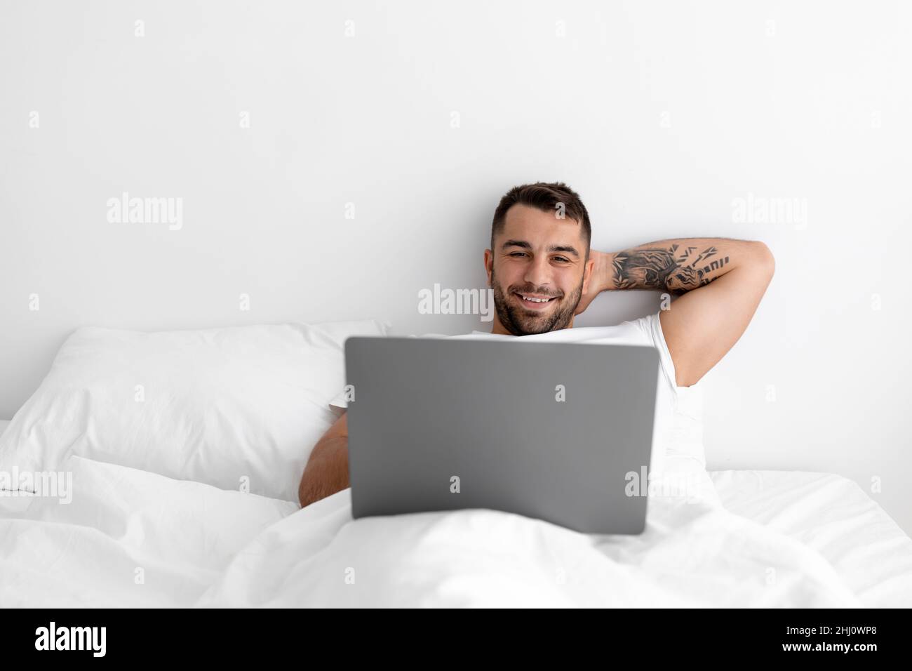 Satisfied relaxed millennial european muscular man lies on white bed ...