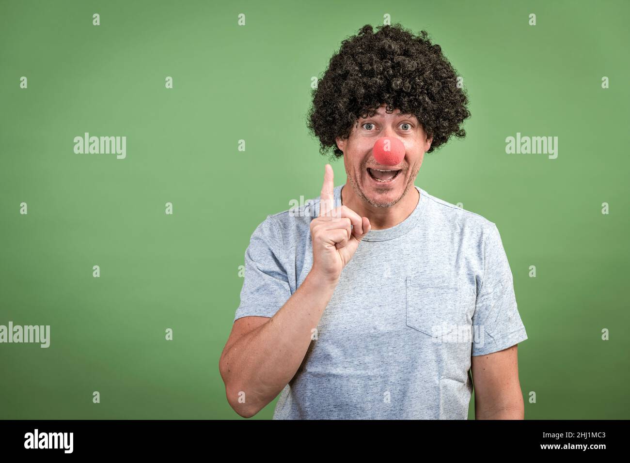 Man with black wig and red clown nose posing in front of green ...