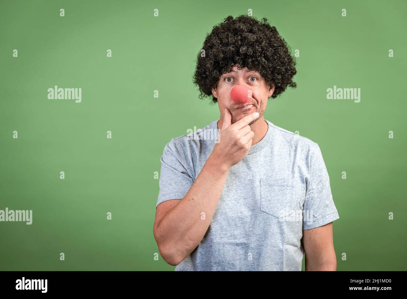 Man with black wig and red clown nose posing in front of green ...