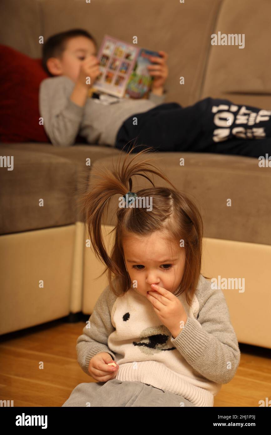 One little kid boy reading at home on the couch while his sister eats ...