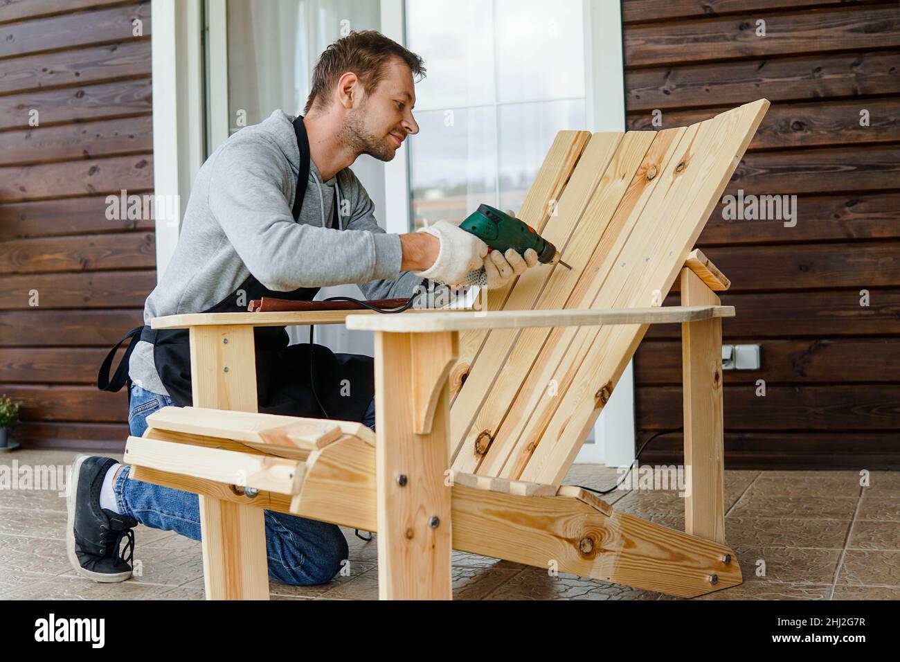 Young carpenter assembling wooden adirondack chair with electric ...