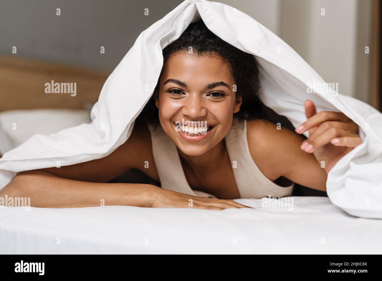 Happy young african woman laying hiding under a blanket on a bed at ...